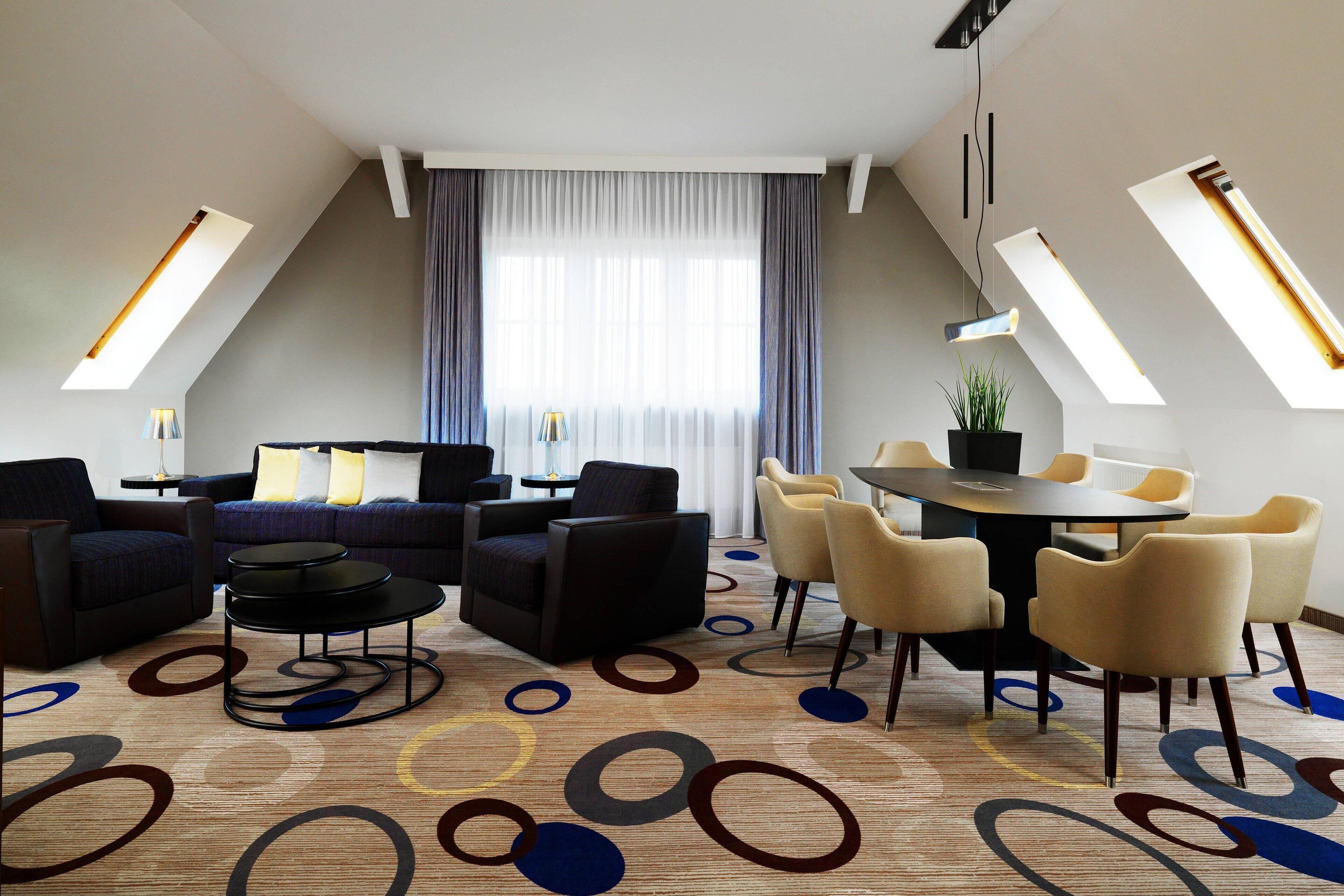 Quarto de hotel moderno com sofá em seção, mesa de jantar redonda com cadeiras, pequena mesa de centro e um vaso de plantas.
