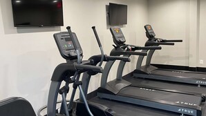 Três equipamentos de exercício com mostradores digitais, uma televisão fixada na parede e uma parede lisa.