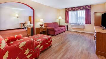 Quarto de hotel com sofá vermelho, poltrona combinando, escrivaninha de madeira com abajur, televisão e janela com cortinas.