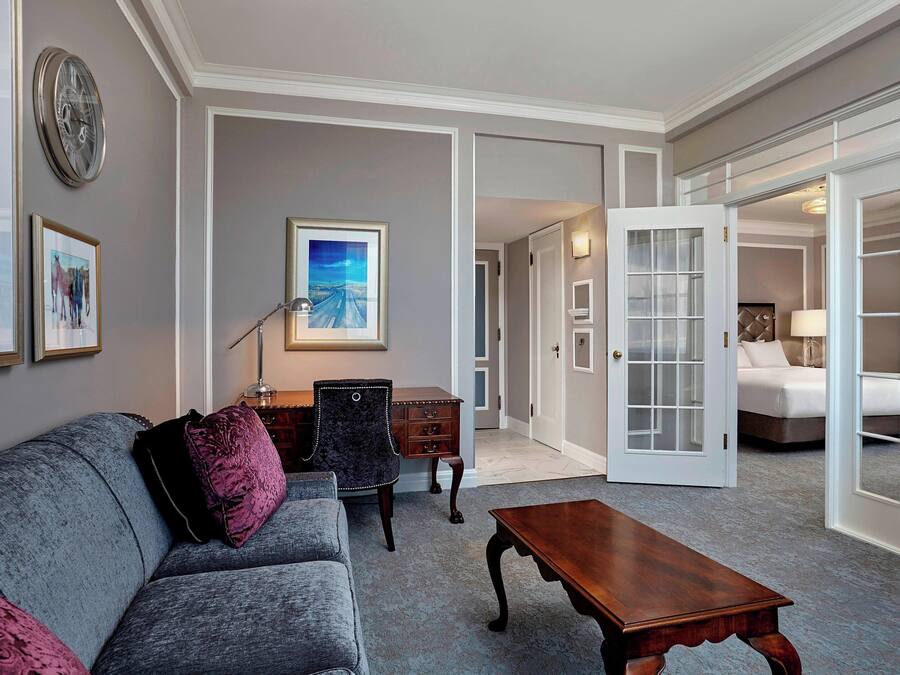 Sala de estar com sofá cinza, mesa de centro de madeira e escrivaninha com luminária. Há uma porta que dá para um quarto com cama e mesa de cabeceira.