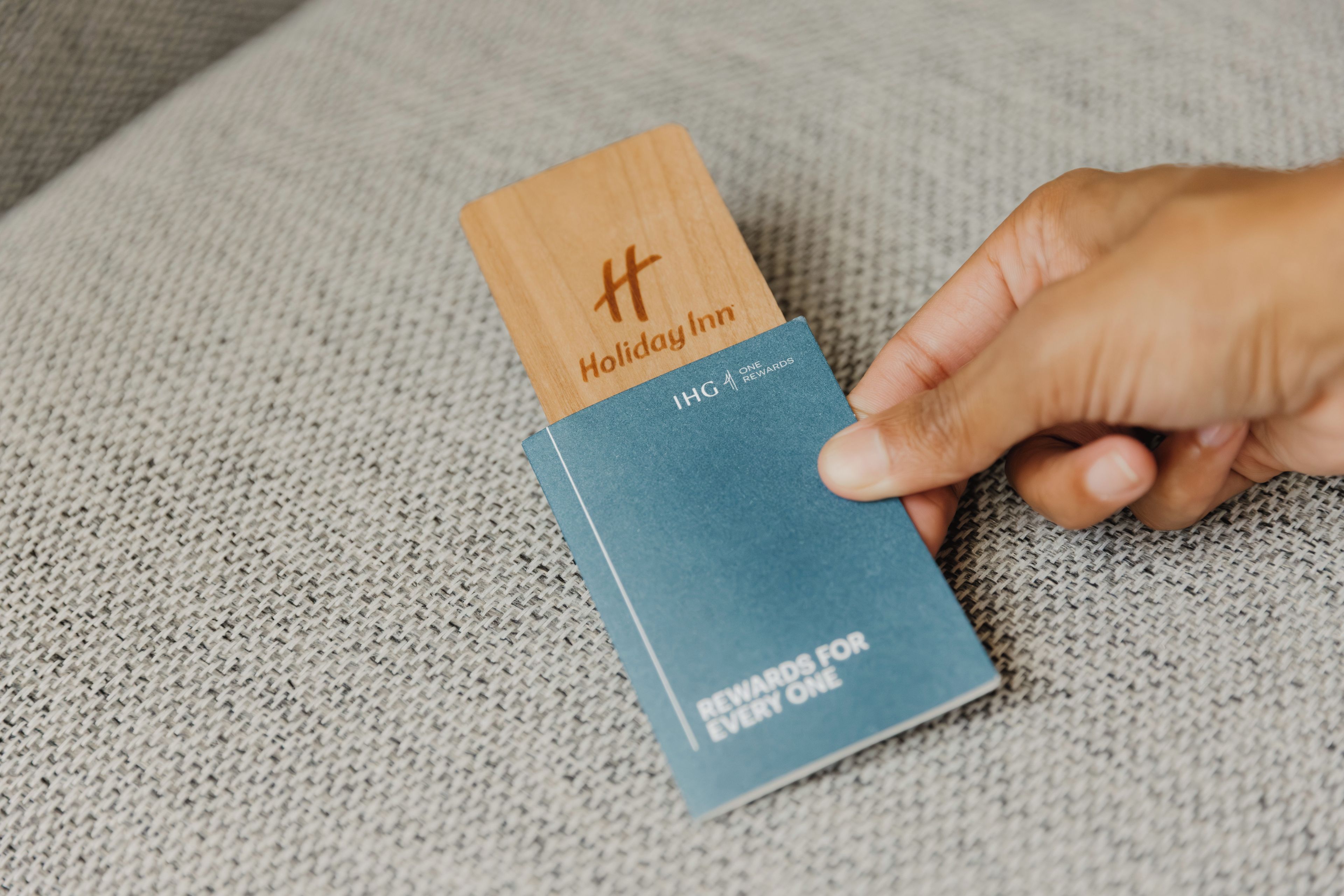 Uma mão segura um cartão de madeira com 'Holiday Inn' e 'ONE REWARDS' visíveis, apoiado em uma superfície de tecido texturizado.