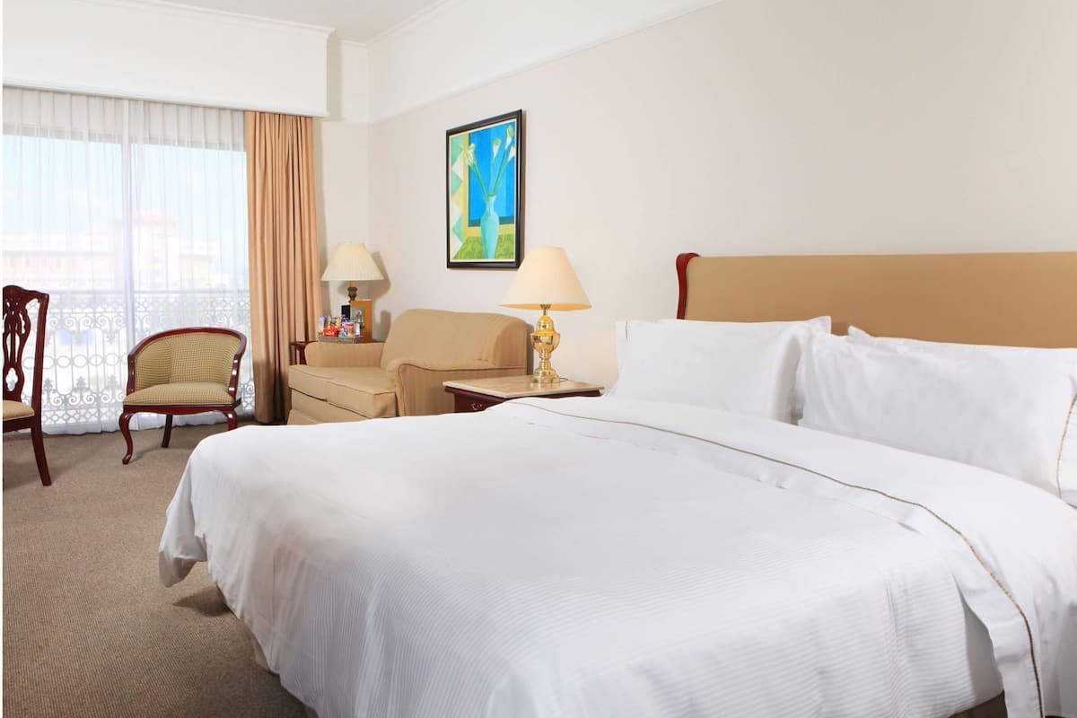 Quarto de hotel com uma cama grande, uma cadeira, uma escrivaninha, um abajur e um quadro na parede.