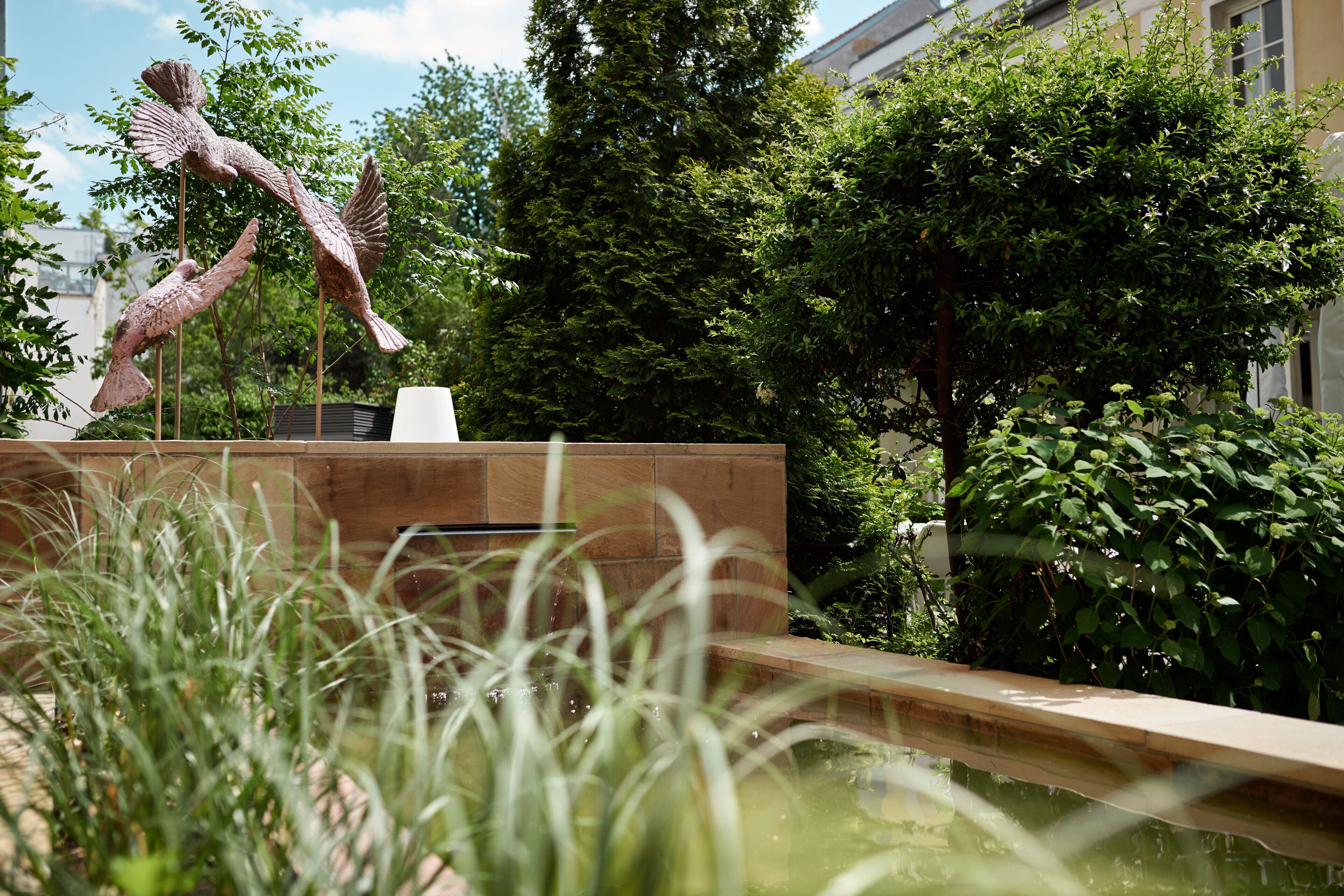 Um jardim com uma escultura de duas aves, um banco e um elemento aquático.