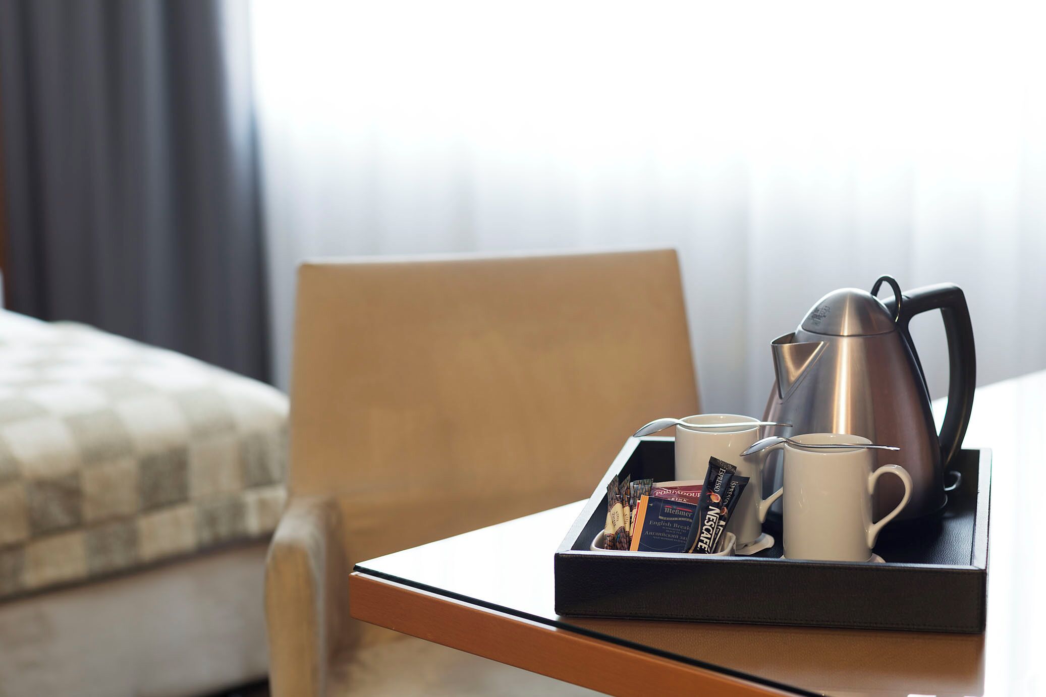 Quarto de hotel com uma cama bem arrumada, uma cadeira de madeira e um tabuleiro com uma cafeteira, duas chávenas e algumas barras de chocolate.