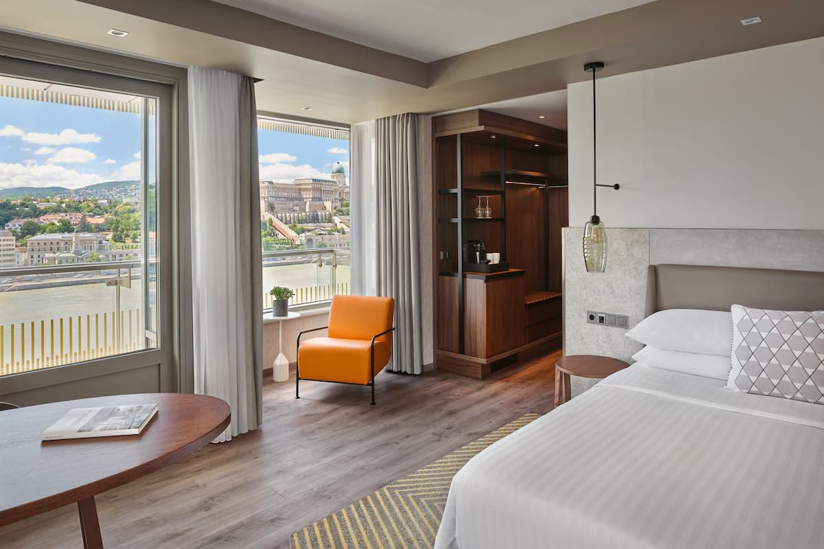Quarto de hotel com uma cama grande, uma escrivaninha de madeira, uma cadeira, uma mesinha e vista para a paisagem urbana através de grandes janelas.