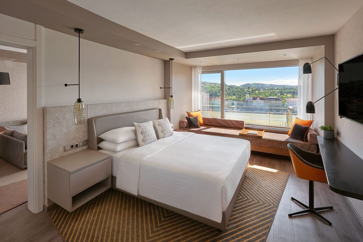 Quarto de hotel moderno com uma cama grande, uma escrivaninha e um sofá. Há uma janela ampla com vista para a cidade.