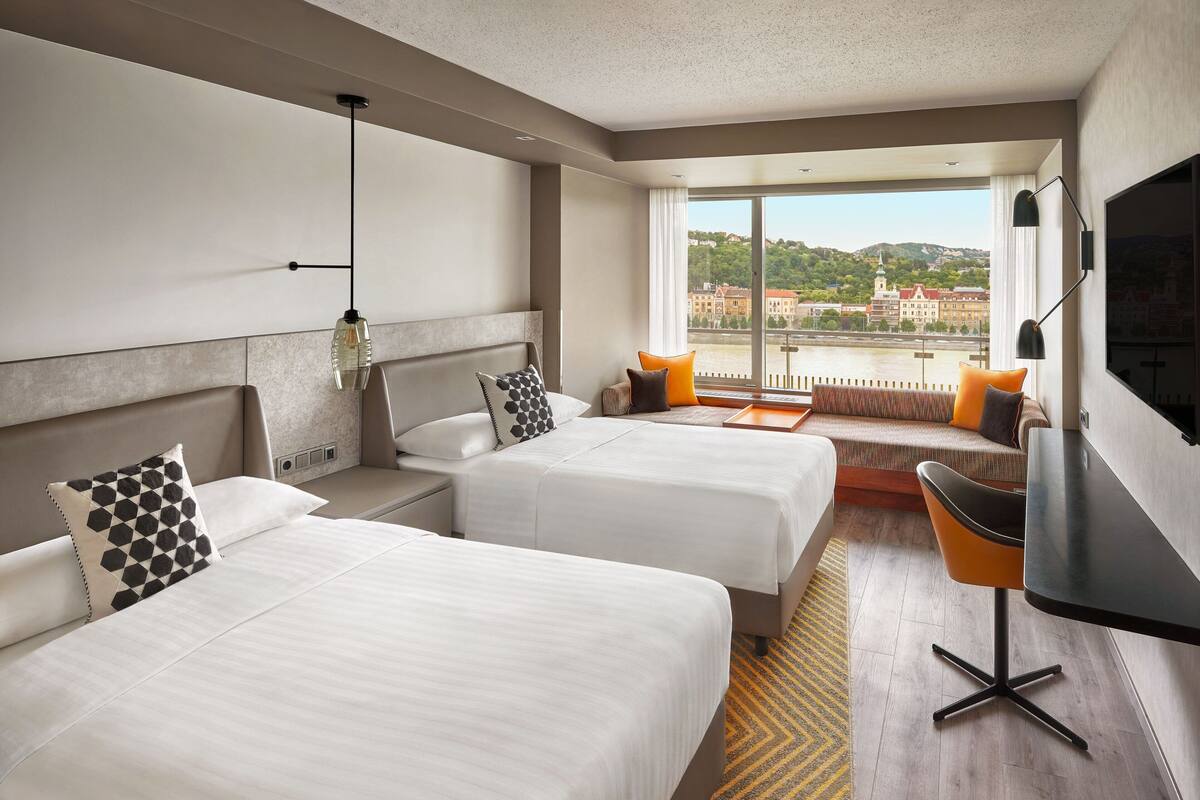 Quarto de hotel com duas camas, um sofá, uma TV e vista para um rio e edifícios.