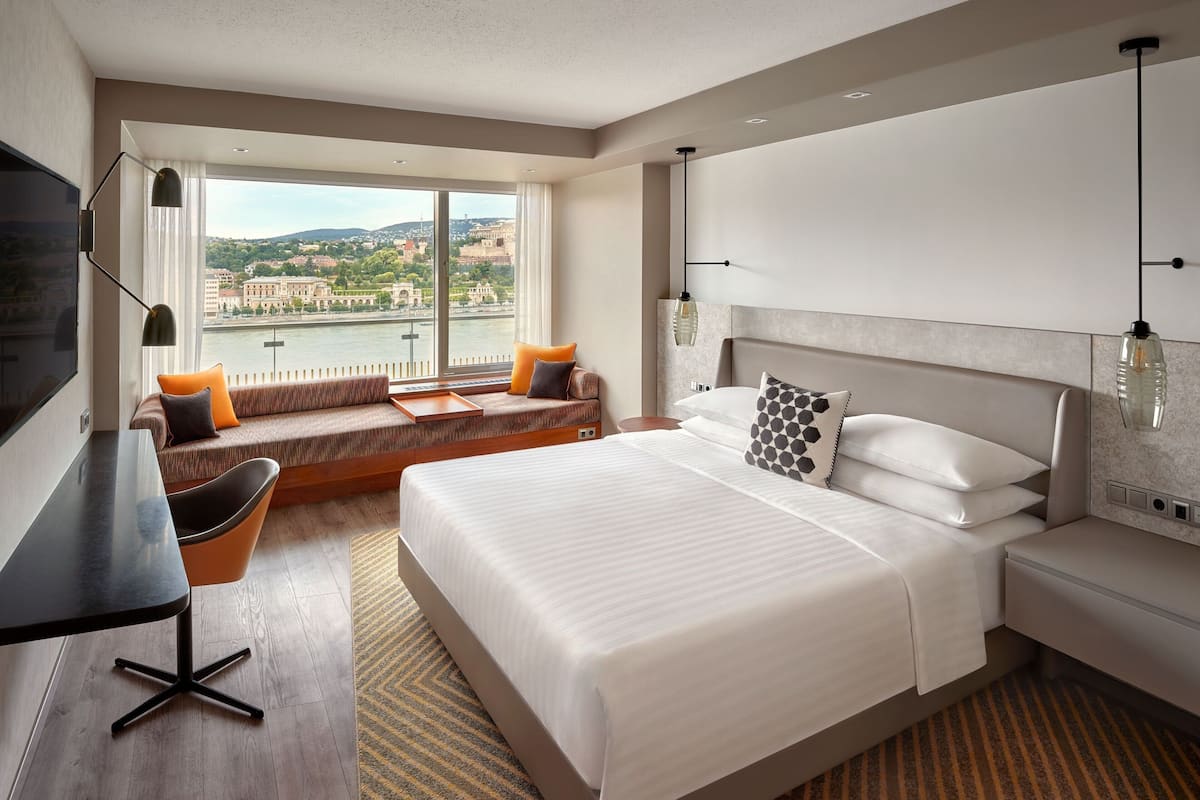 Quarto de hotel moderno com cama grande, escrivaninha, sofá e vista para um rio e horizonte urbano.