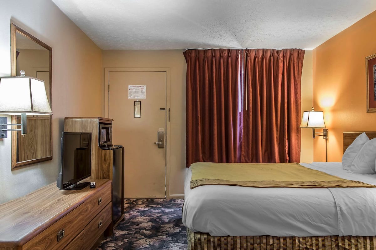 Quarto de hotel com uma cama, uma televisão sobre uma cômoda de madeira, uma porta com um aviso e uma janela com cortinas vermelhas.