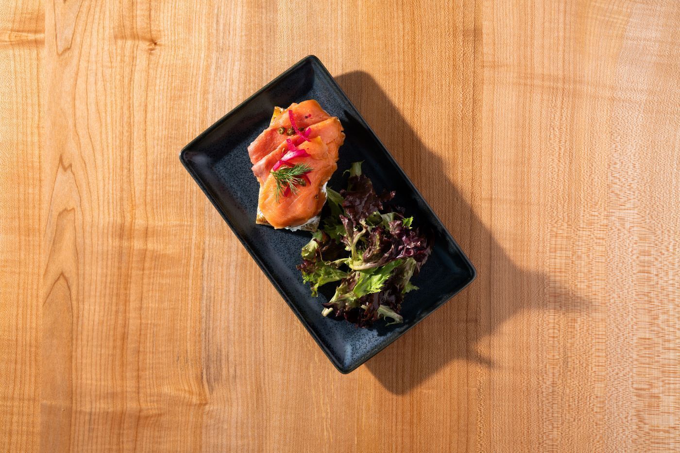 Prato preto retangular com salmão e salada verde mista sobre uma mesa de madeira.