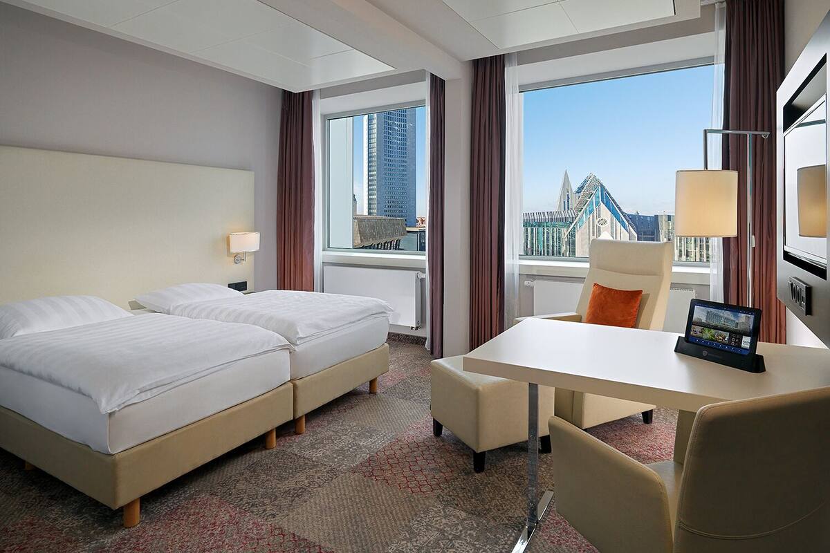 Quarto de hotel com duas camas, uma mesinha, uma cadeira, televisão e janela com vista para a cidade.