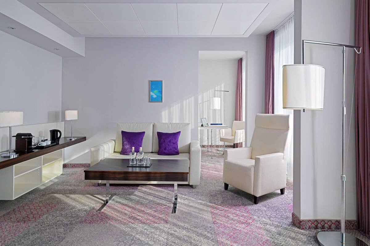 Quarto de hotel moderno com sofá, poltrona, mesa de centro e escrivaninha com abajur.