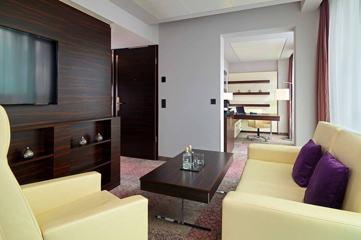 Quarto de hotel moderno com televisão de tela plana, um armário de madeira escura com prateleiras, um sofá cor creme com almofadas roxas e uma pequena mesa de centro.