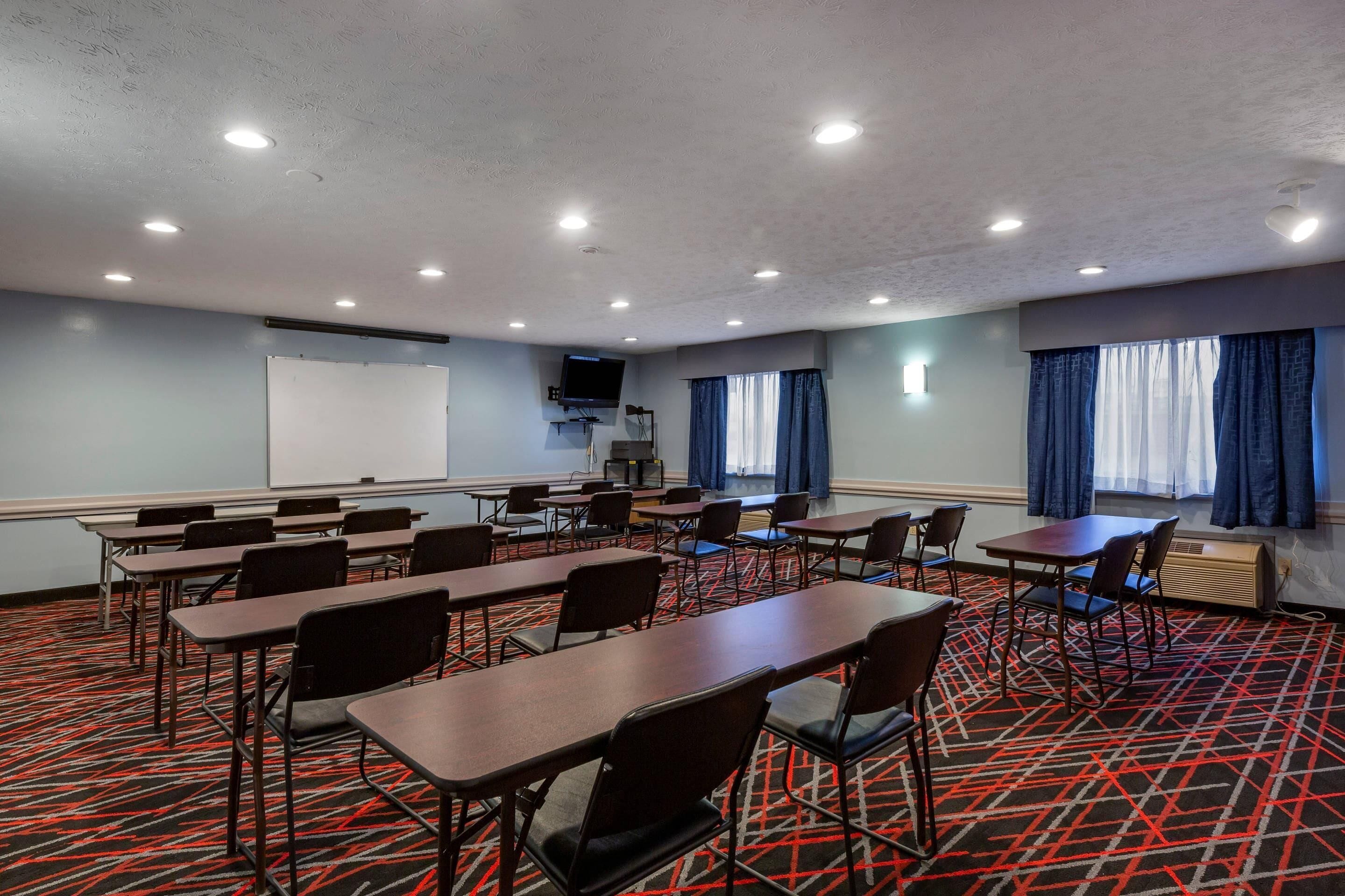 Uma sala de conferências com mesas, cadeiras, um quadro branco, uma tela de projetor e uma televisão.