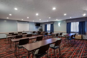 Uma sala de conferências com mesas, cadeiras, um quadro branco, uma tela de projetor e uma televisão.