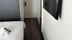 Quarto de hotel com uma cama, televisão fixada na parede, porta com uma placa e chão coberto de carpete.