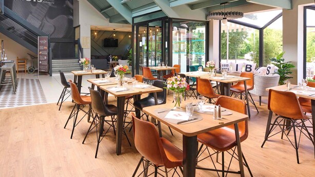 Restaurante com mesas de madeira e cadeiras laranja, uma escada e uma grande janela com vegetação lá fora.