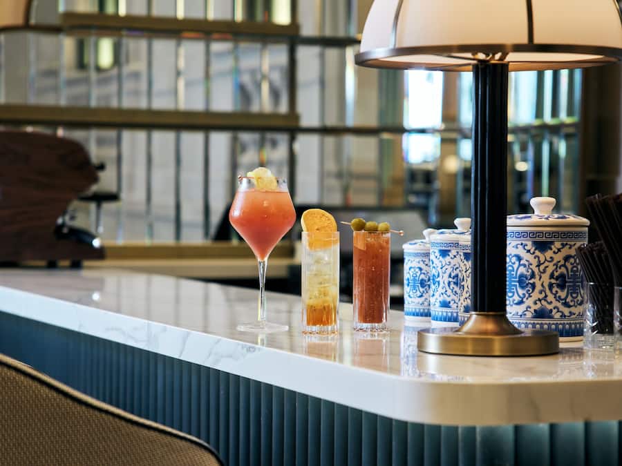 Uma área de bar com bancada de mármore, uma luminária branca e três bebidas.