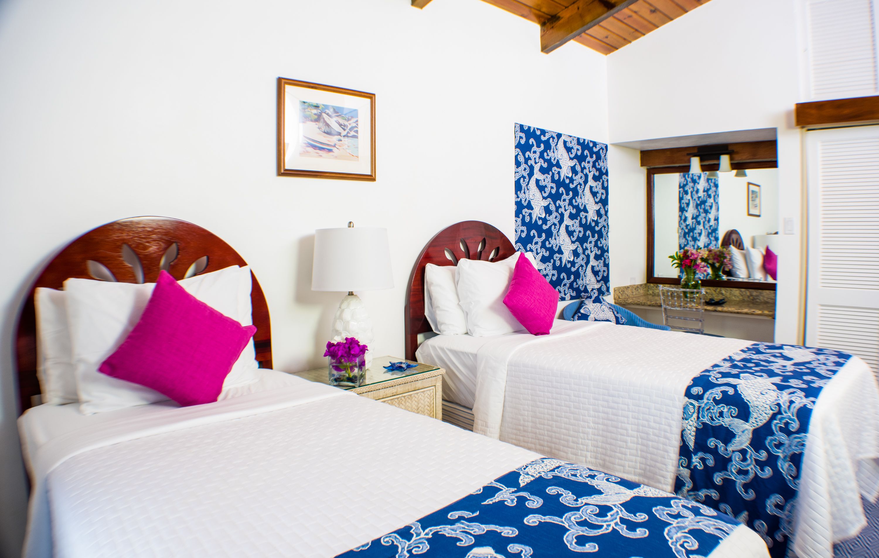 Quarto de hotel com duas camas, cada uma com cabeceira de madeira, roupa de cama branca e colcha estampada em azul e branco. Há uma mesa de cabeceira com uma luminária e um vaso de flores. Um quadro emoldurado está pendurado na parede, e um espelho reflete parte do quarto.