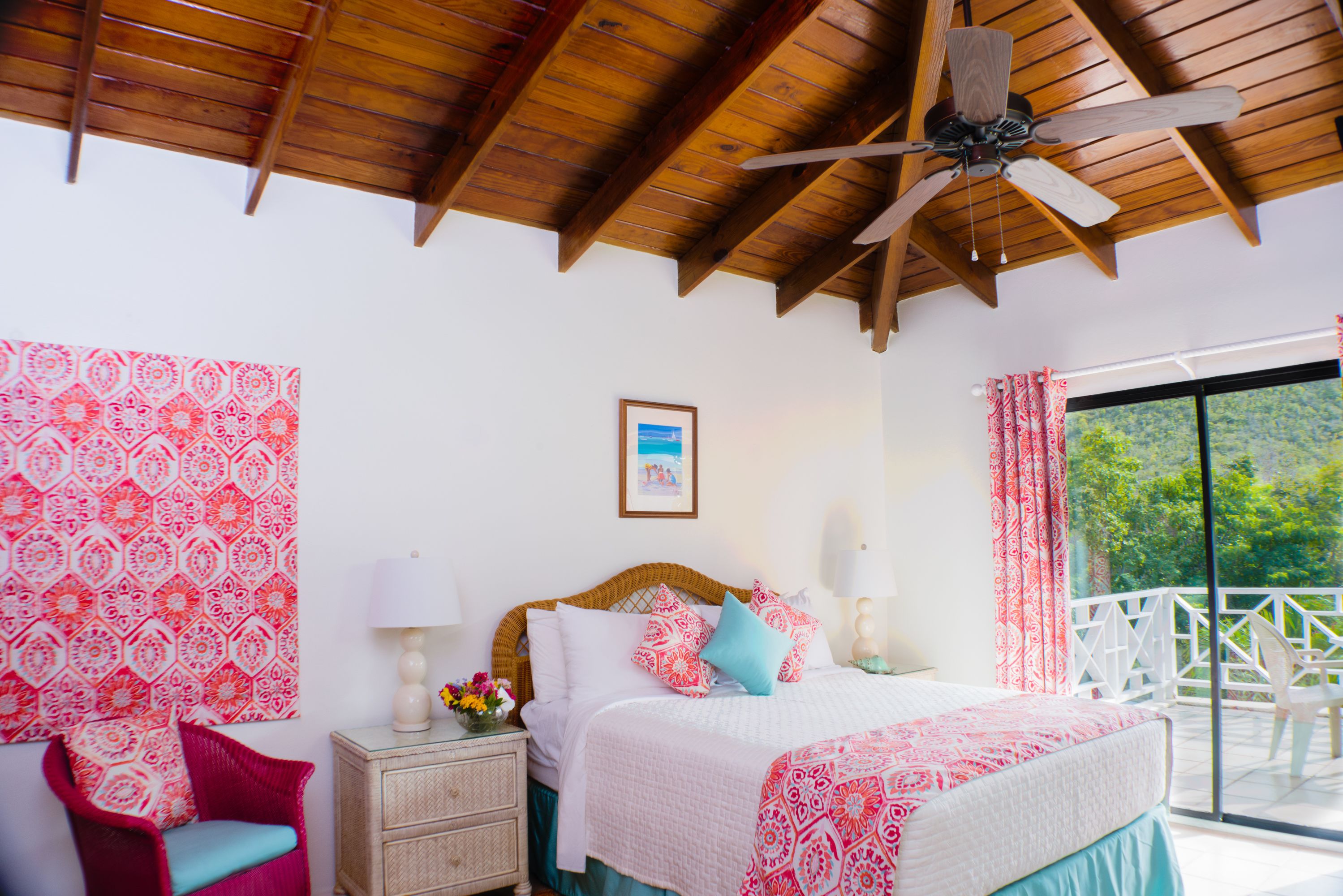 Quarto com cama, mesas de cabeceira, poltrona, ventoinha no teto e janela com vista para vegetação.