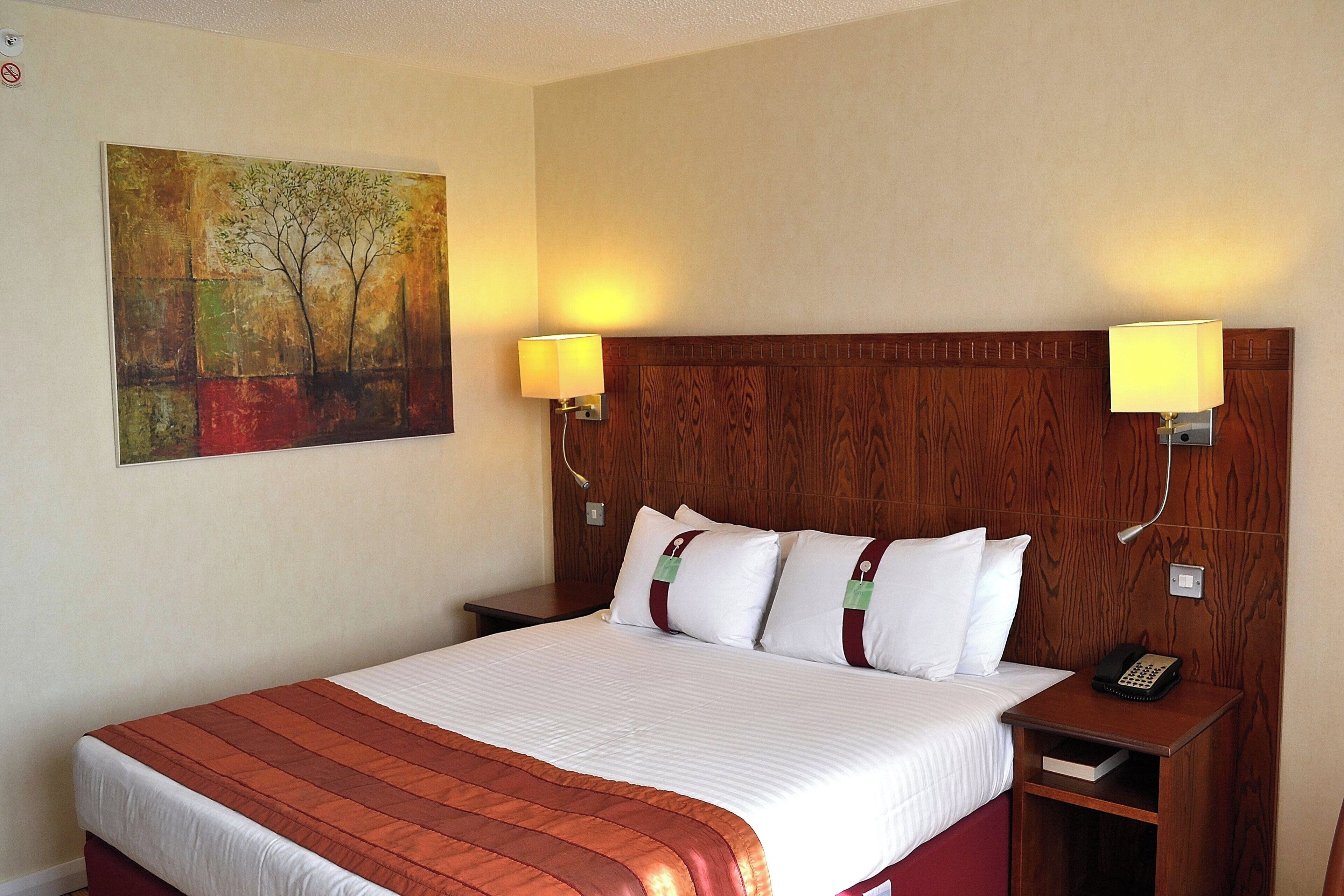 Quarto de hotel com cama, duas luminárias de cabeceira, um quadro na parede e mesa de cabeceira com telefone.