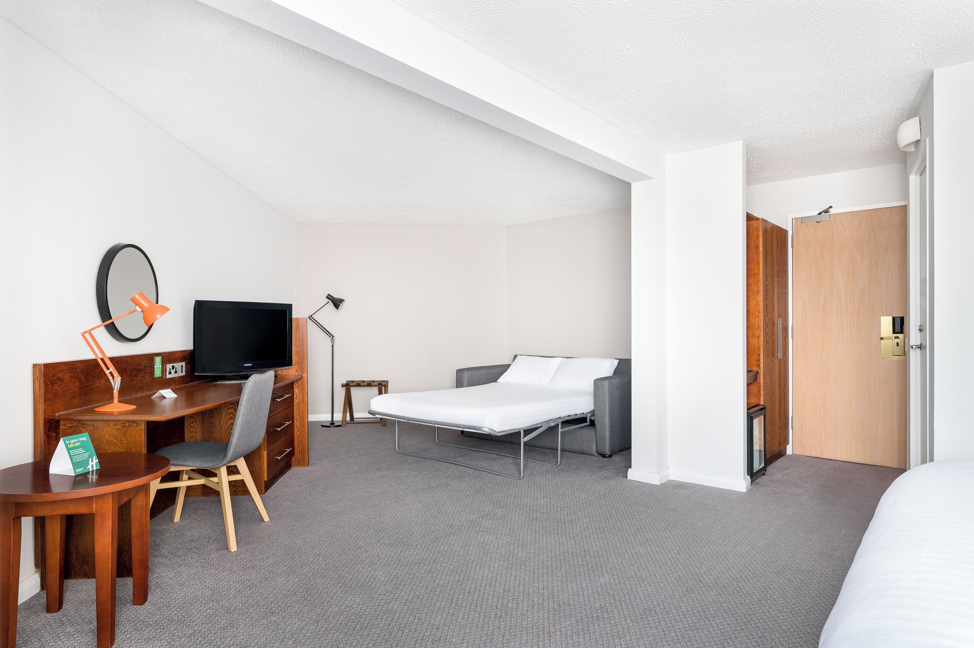 Quarto de hotel com escrivaninha, cadeira, televisão, cama e guarda-roupa.