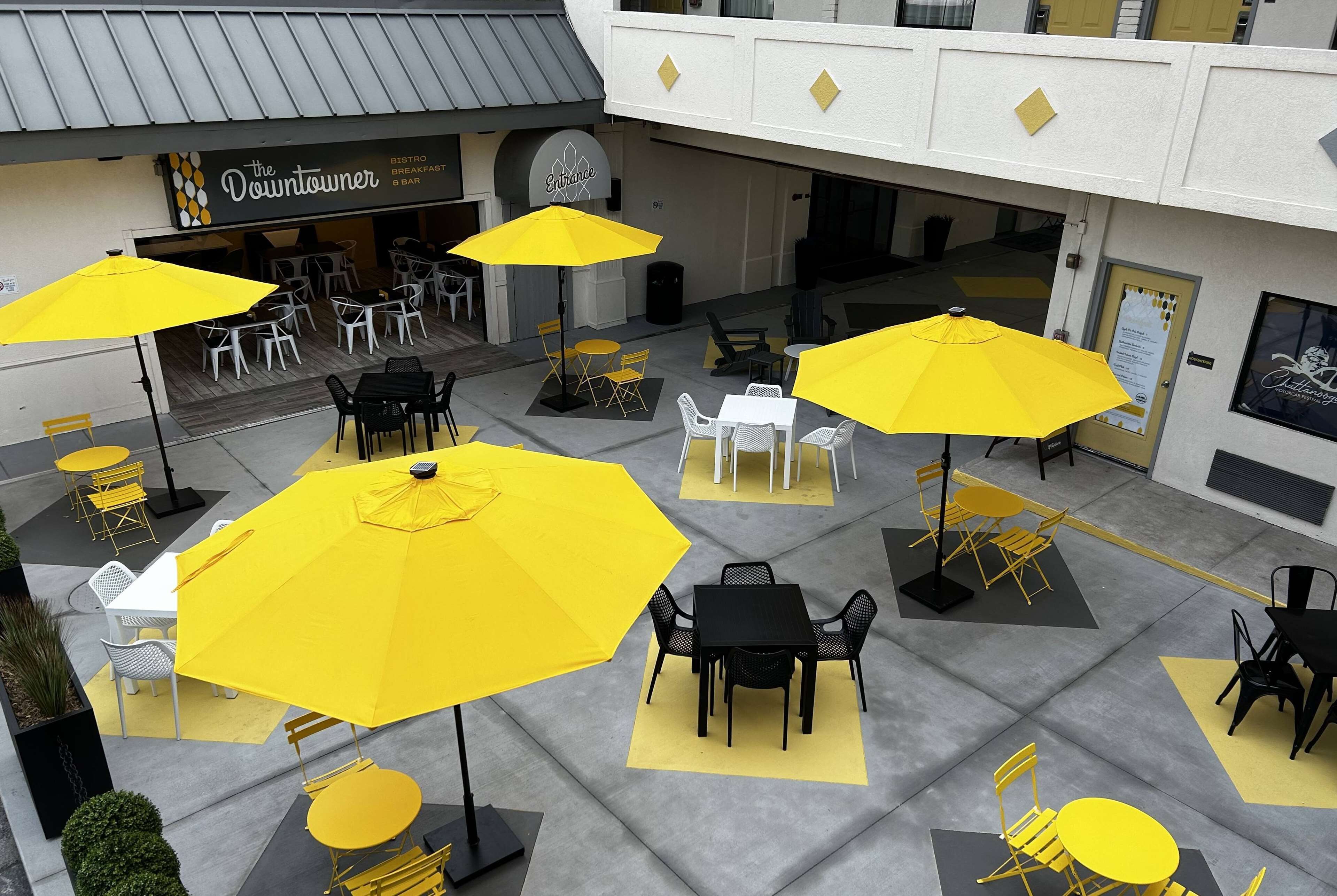 Uma área de pátio ao ar livre com guarda-sóis amarelos e mesas, um letreiro do restaurante 'The Downtowner' e um edifício branco ao fundo.
