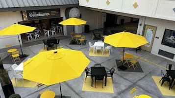 Uma área de pátio ao ar livre com guarda-sóis amarelos e mesas, um letreiro do restaurante 'The Downtowner' e um edifício branco ao fundo.
