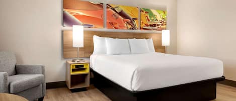 Quarto de hotel moderno com uma cama grande, uma poltrona cinza, uma mesinha e uma obra de arte pendurada na parede.