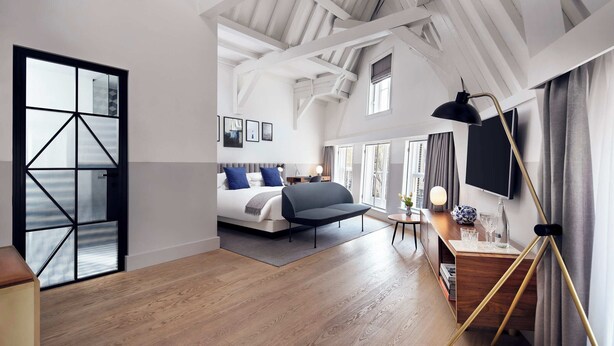 Quarto de hotel moderno com uma cama grande, uma mesa com abajur e uma televisão na parede.