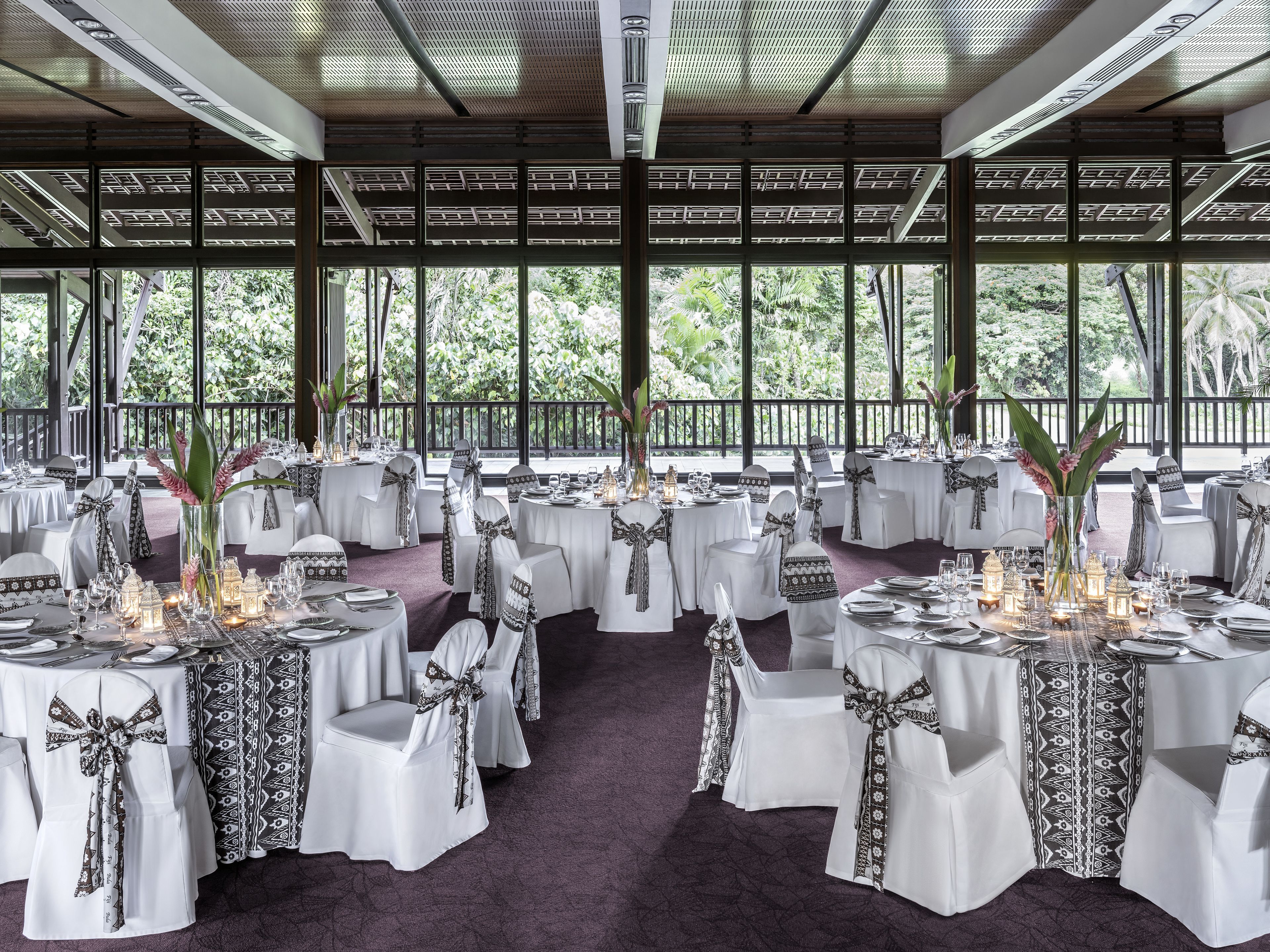 Uma sala de banquetes com mesas redondas, cadeiras brancas e capas estampadas. As mesas estão montadas com pratos, copos e velas.