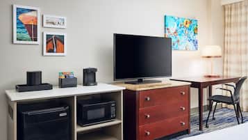 Quarto de hotel com TV de tela plana, cafeteira, micro-ondas, mini-geladeira, escrivaninha com luminária e obra de arte na parede.