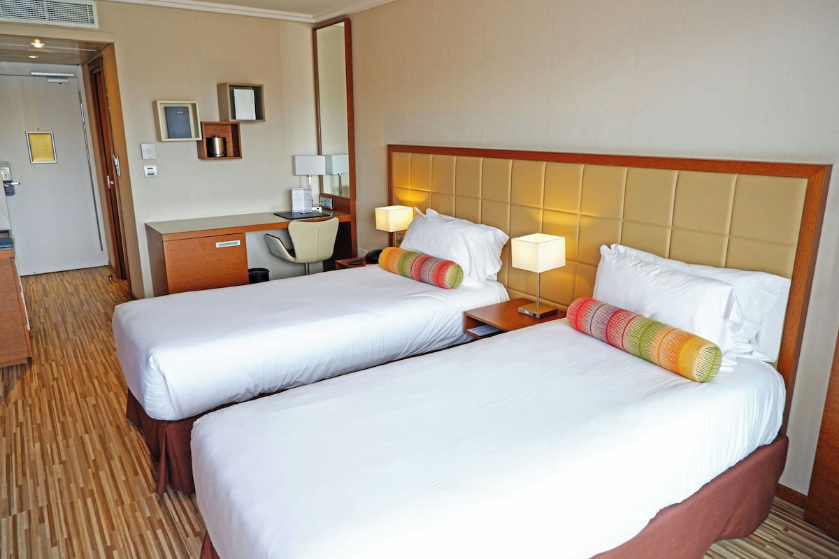 Quarto de hotel com duas camas de solteiro, uma escrivaninha, uma cadeira, um espelho e mesa de cabeceira com luminárias.