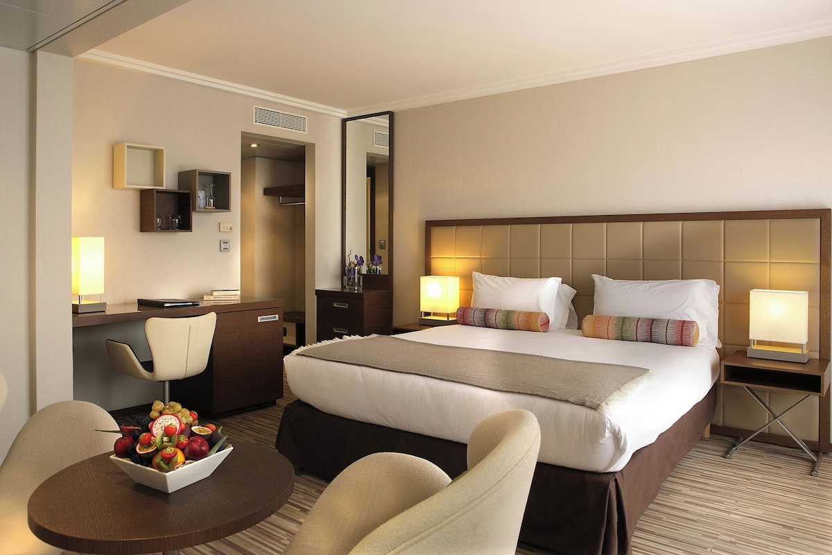 Quarto de hotel com cama, abajures, escrivaninha com cadeira, espelho e uma cesta de frutas.