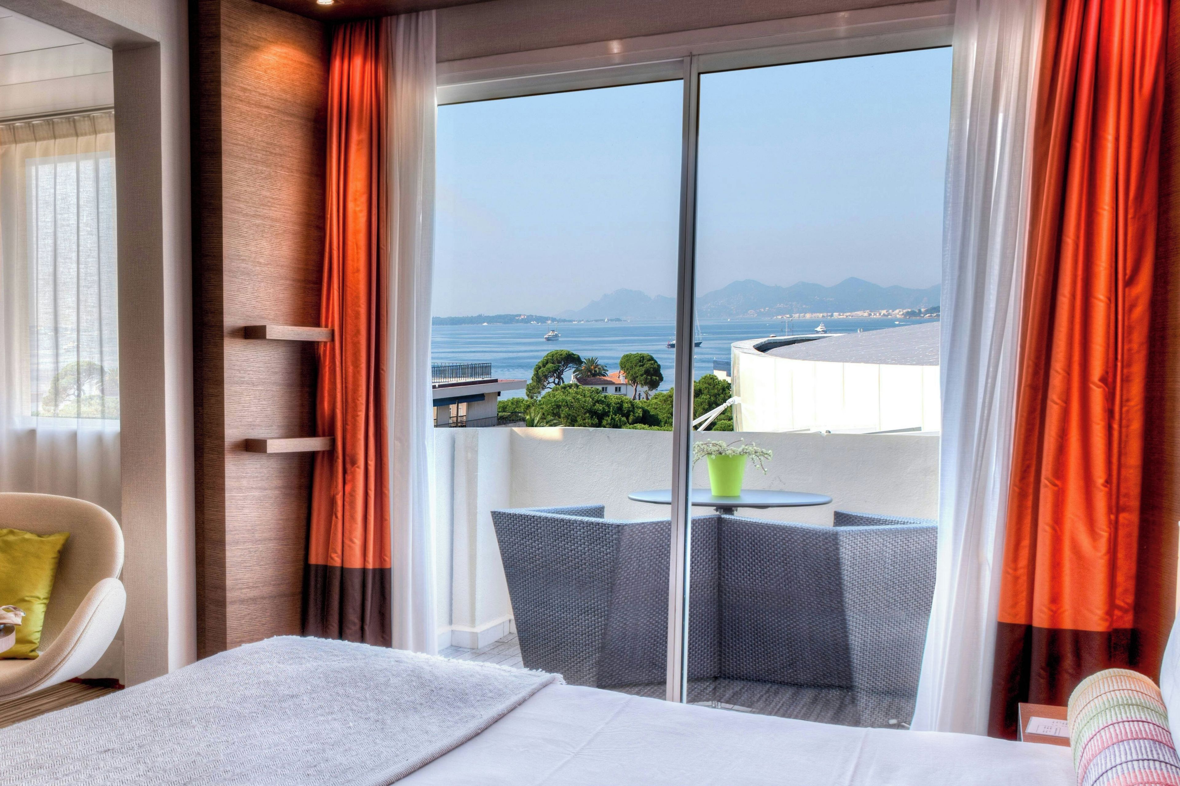 Quarto de hotel com cama, uma cadeira, mesa pequena e varanda com vista para o mar e montanhas.