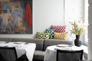 Uma sala de jantar com um sofá e cadeiras, uma mesa posta com pratos e talheres, um quadro na parede e um vaso com flores junto à janela.