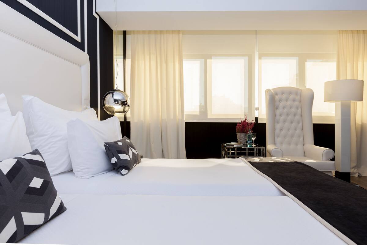 Quarto de hotel moderno com uma cama grande, um espaço de estar com uma poltrona e uma mesa, e uma janela ampla com cortinas.