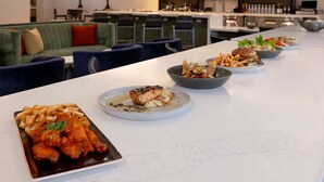 Uma sala de jantar com uma mesa comprida exibindo diversos pratos, um espaço de bar com bancos, e uma área de estar com sofá e cadeiras.