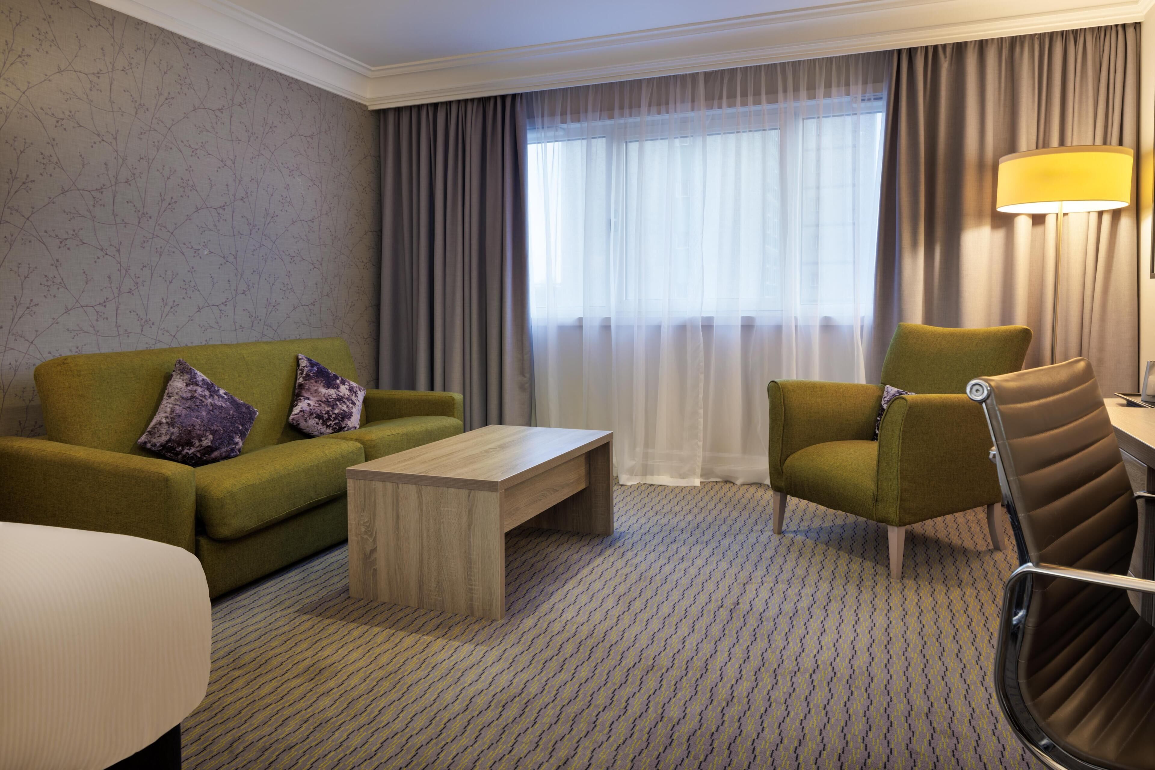 Quarto de hotel com sofá verde, mesa de centro de madeira, poltrona marrom, escrivaninha e luminária.