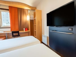 Quarto de hotel com cama, escrivaninha, cadeira, televisão e guarda-roupa.