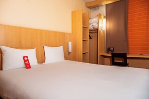 Quarto de hotel com cama, cabeceira de madeira, guarda-roupa e mesa de cabeceira com abajur.