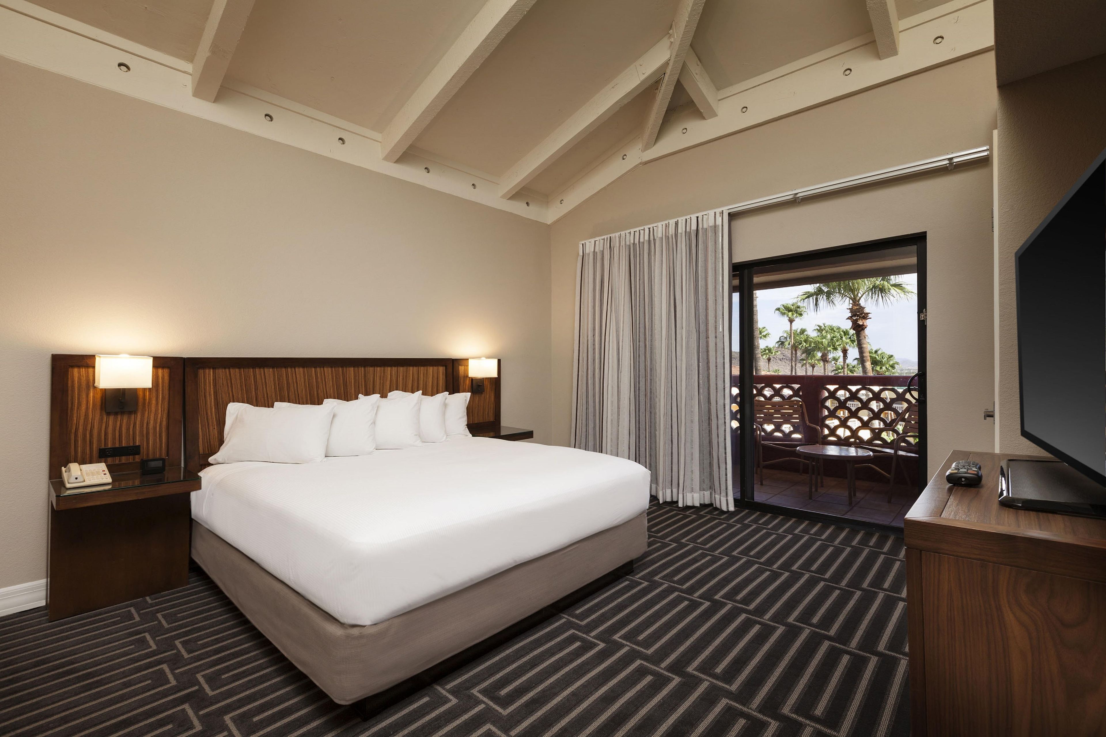 Quarto de hotel com uma cama grande, escrivaninha com telefone, televisão e varanda com vista para palmeiras.