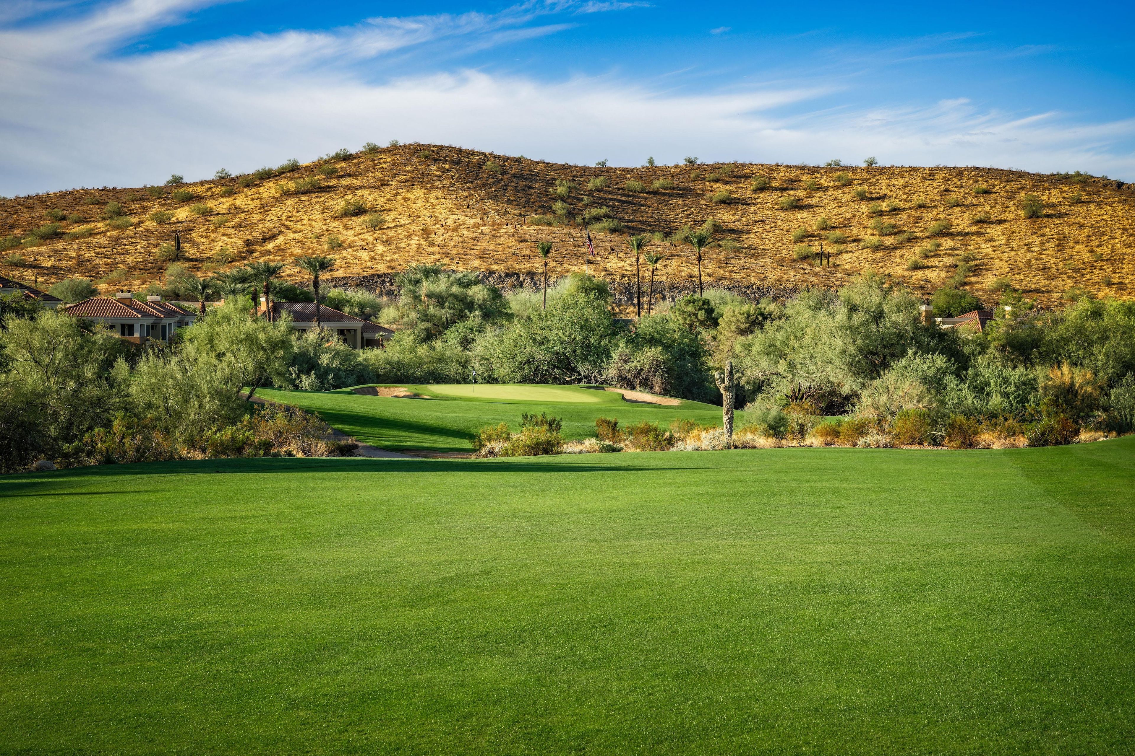Um campo de golfe sob um céu claro, com um green bem cuidado e um cenário montanhoso ao fundo.