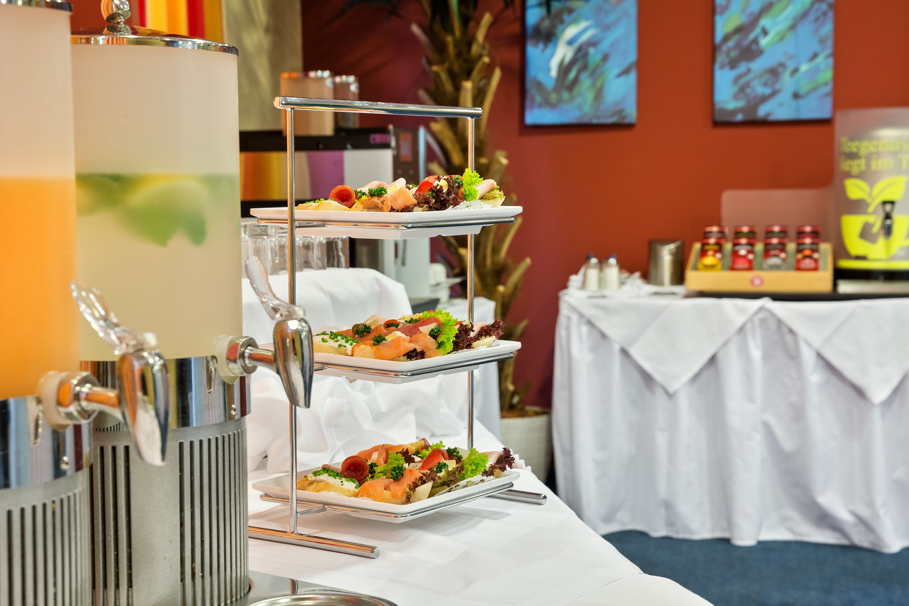 Buffet montado com bandejas de servir em três níveis, um dispensador de bebidas e uma mesa com condimentos.