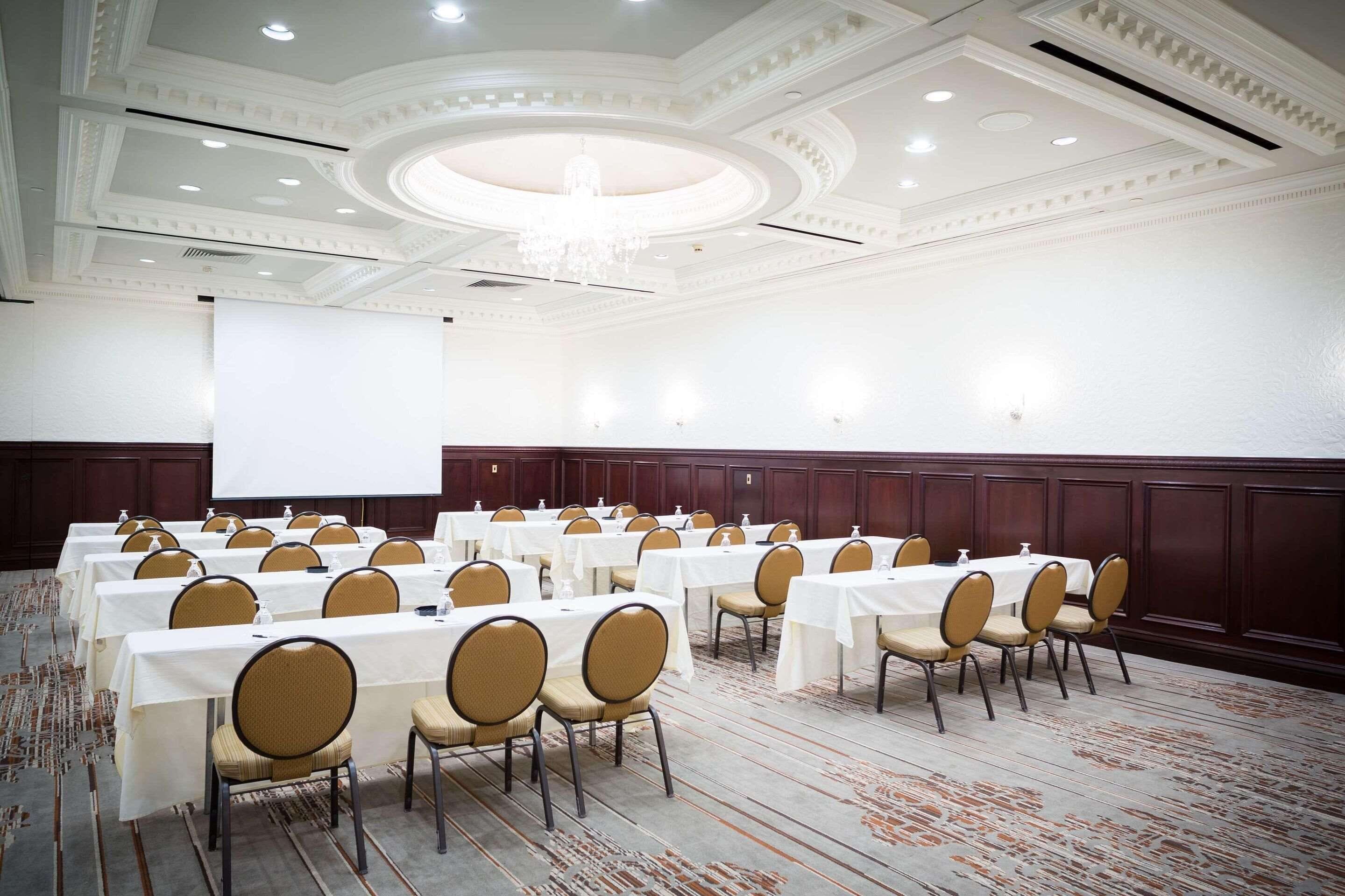 Sala de conferências com várias mesas e cadeiras, uma tela de projetor e um candelabro.