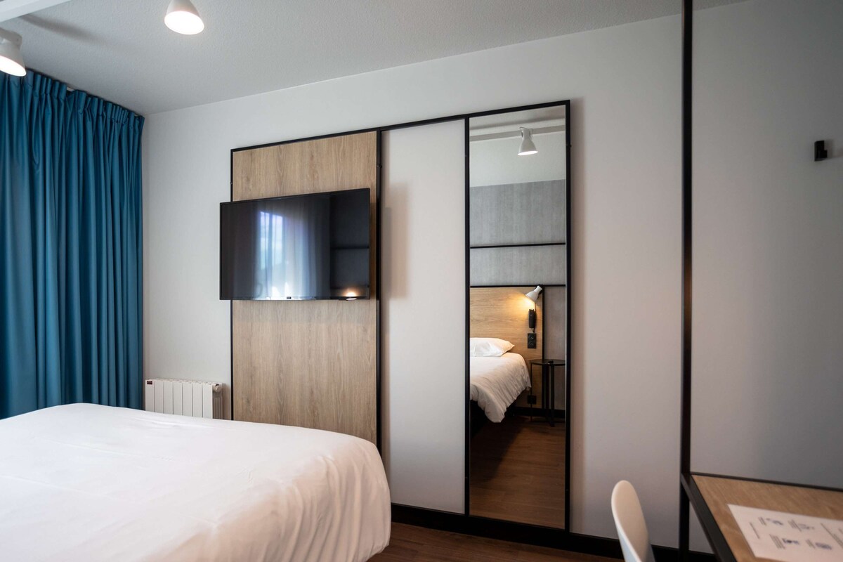 Quarto de hotel com cama, escrivaninha, cadeira, TV presa na parede e um espelho refletindo o quarto.