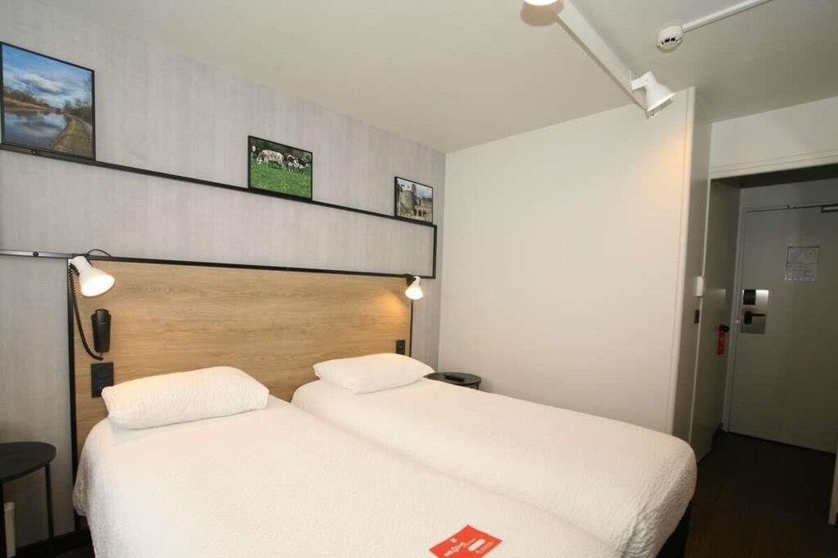 Quarto de hotel com cama, abajures, cabeceira, mesinha e porta.