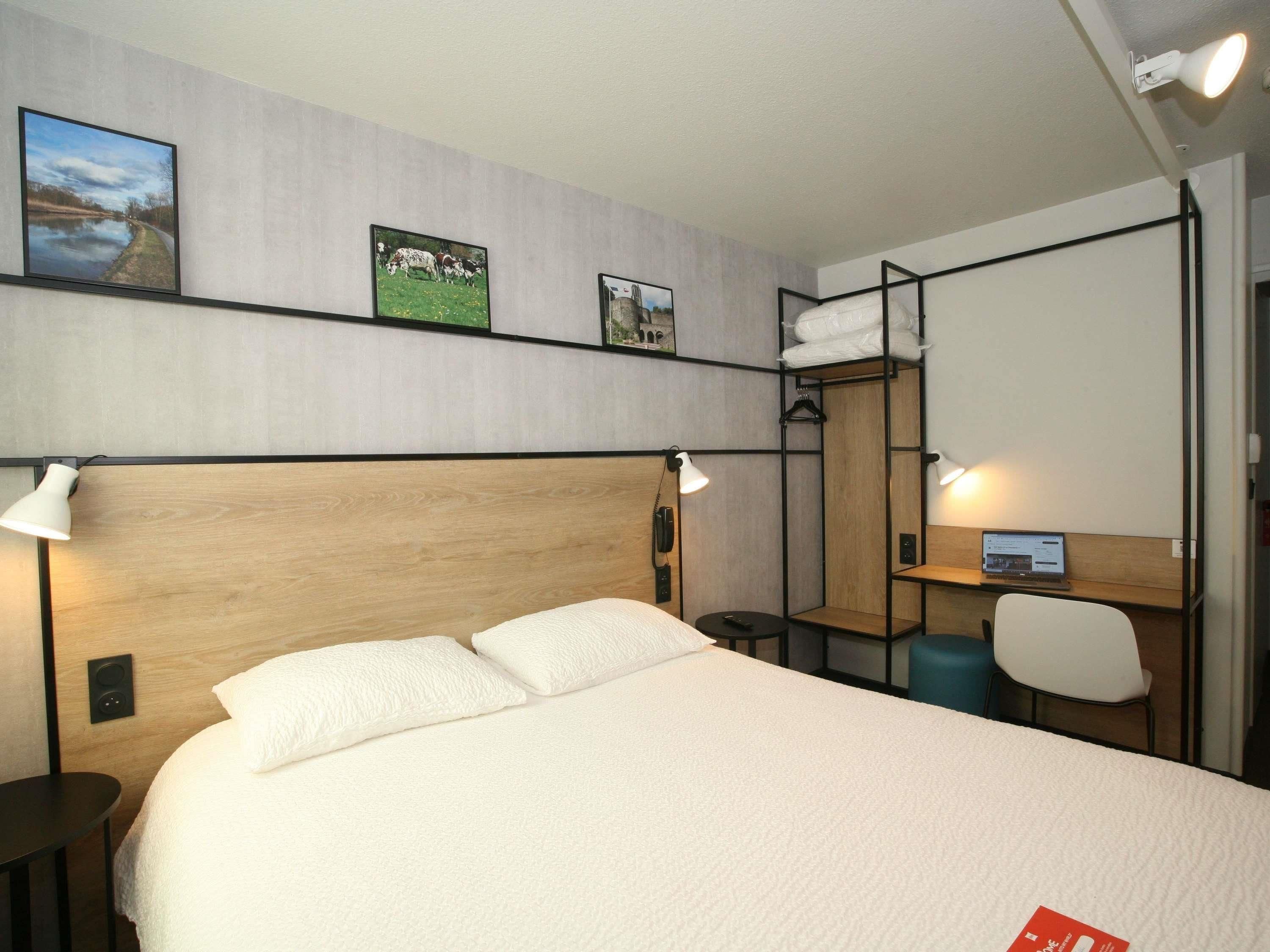 Quarto de hotel com cama, luminárias laterais, escrivaninha com um laptop, cadeira e prateleiras na parede com quadros.