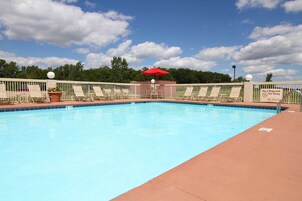 Uma piscina com espreguiçadeiras e um guarda-sol vermelho, cercada por uma grade com uma placa indicando ausência de salva-vidas.