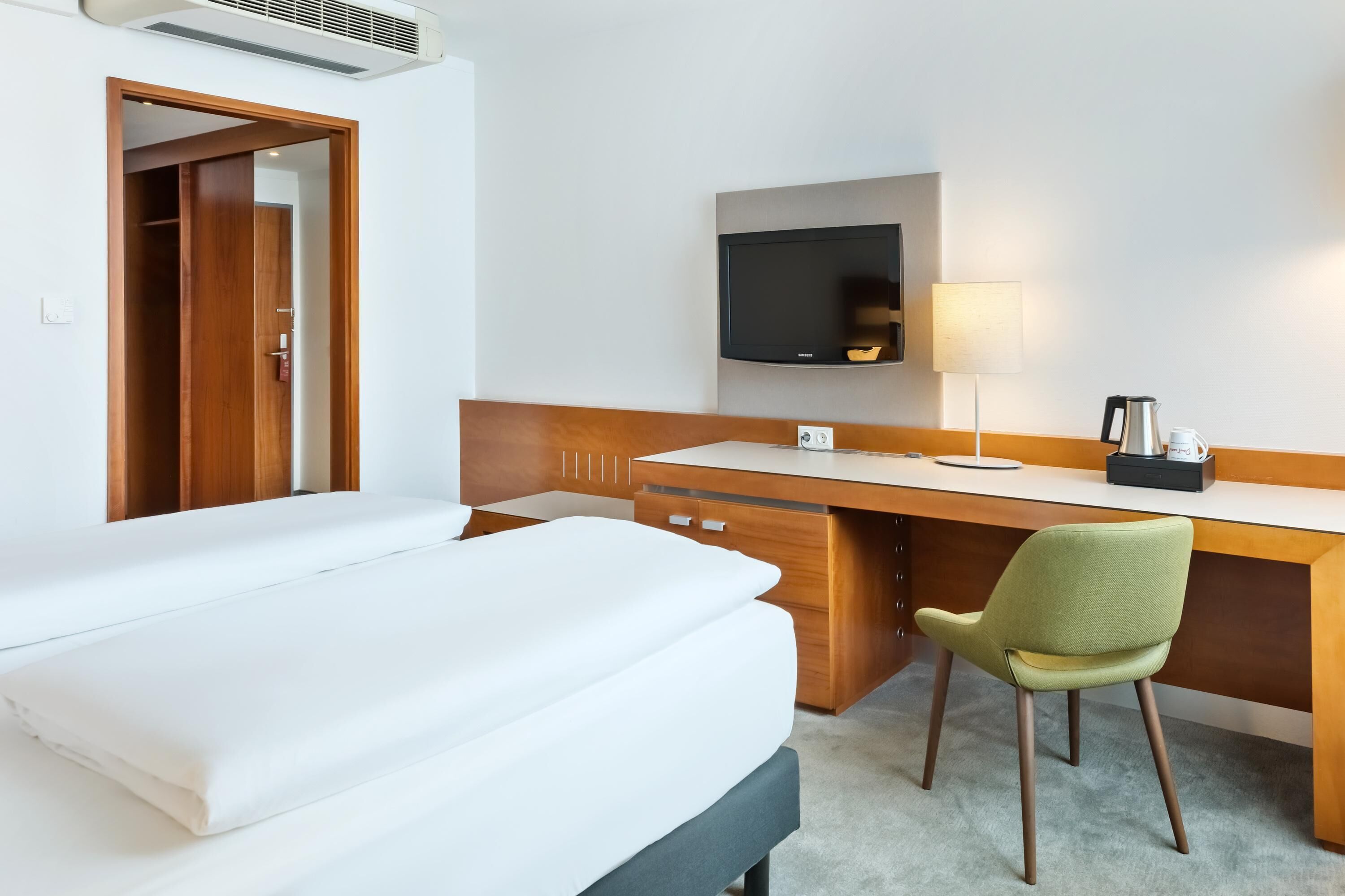 Quarto de hotel com cama, escrivaninha, cadeira, televisão e luminária.
